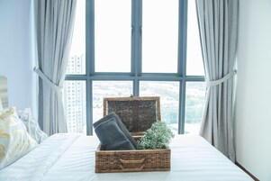 Uma cama com colcha branca, uma cesta de vime com uma toalha cinza e uma pequena planta, além de vista para edifícios através de uma janela com cortinas.