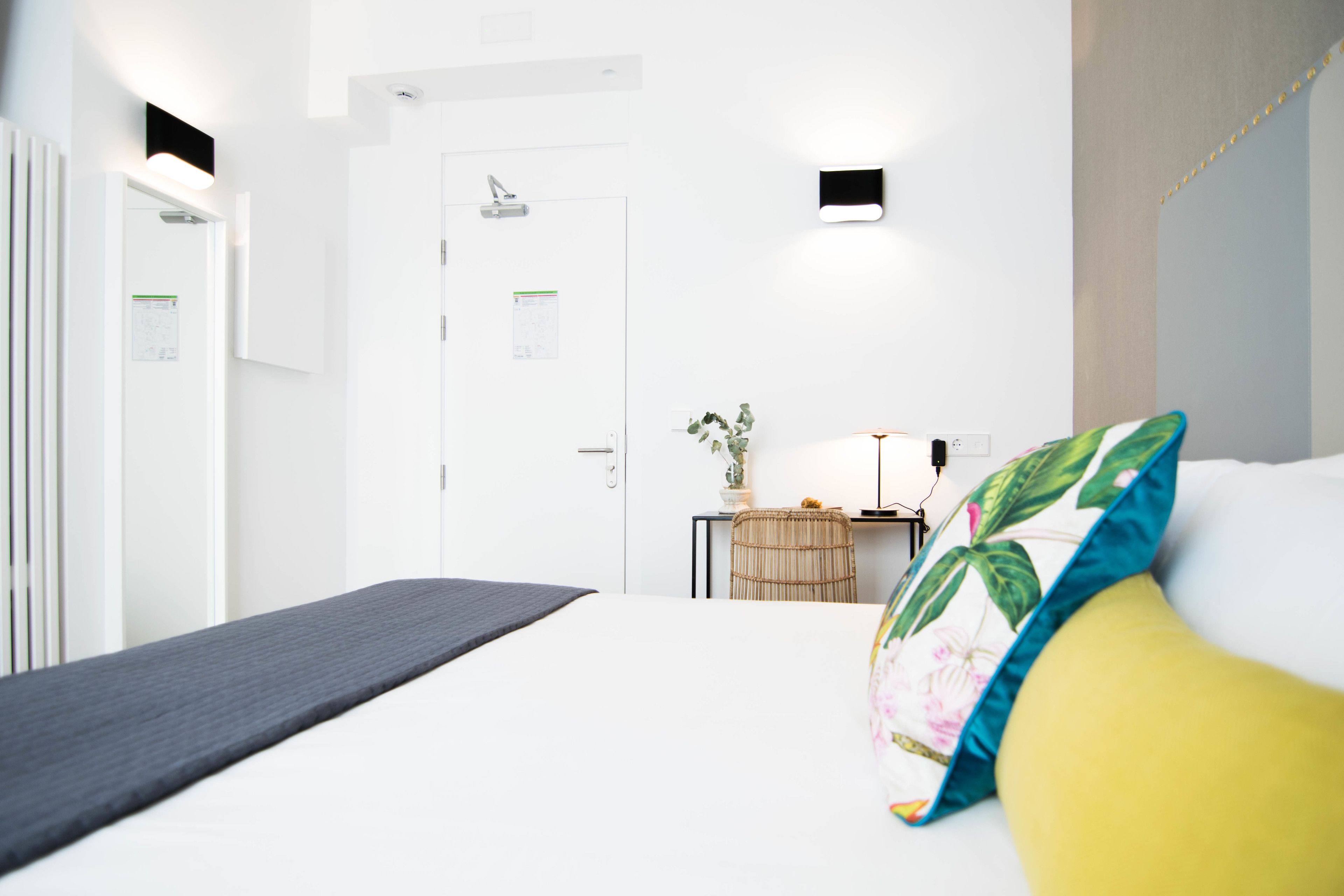 Quarto de hotel moderno com uma cama, mesa de cabeceira com abajur, planta em vaso e porta com um calendário.