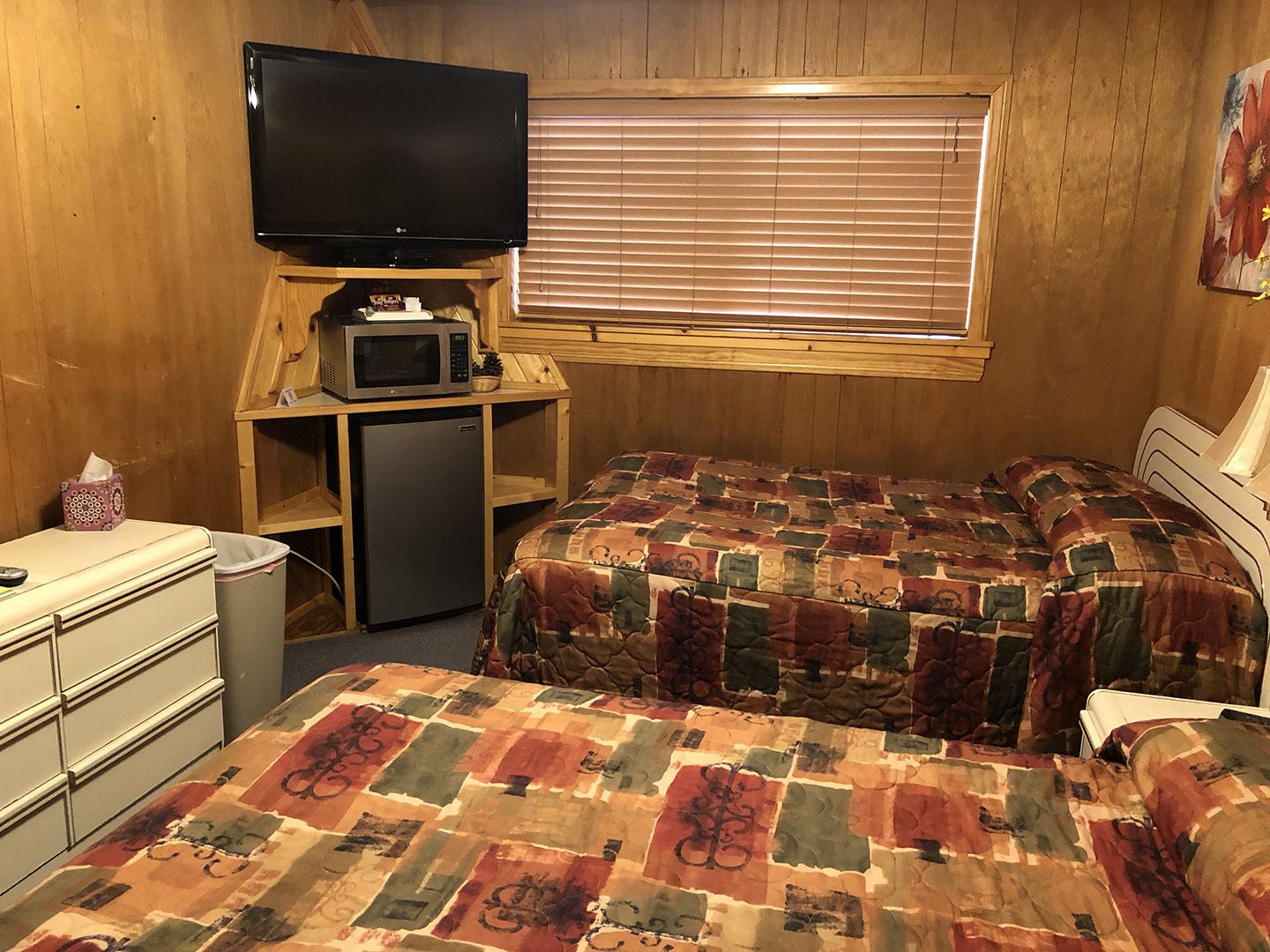 Quarto compacto com uma cama, uma pequena escrivaninha com computador, televisão e uma janela com persianas.