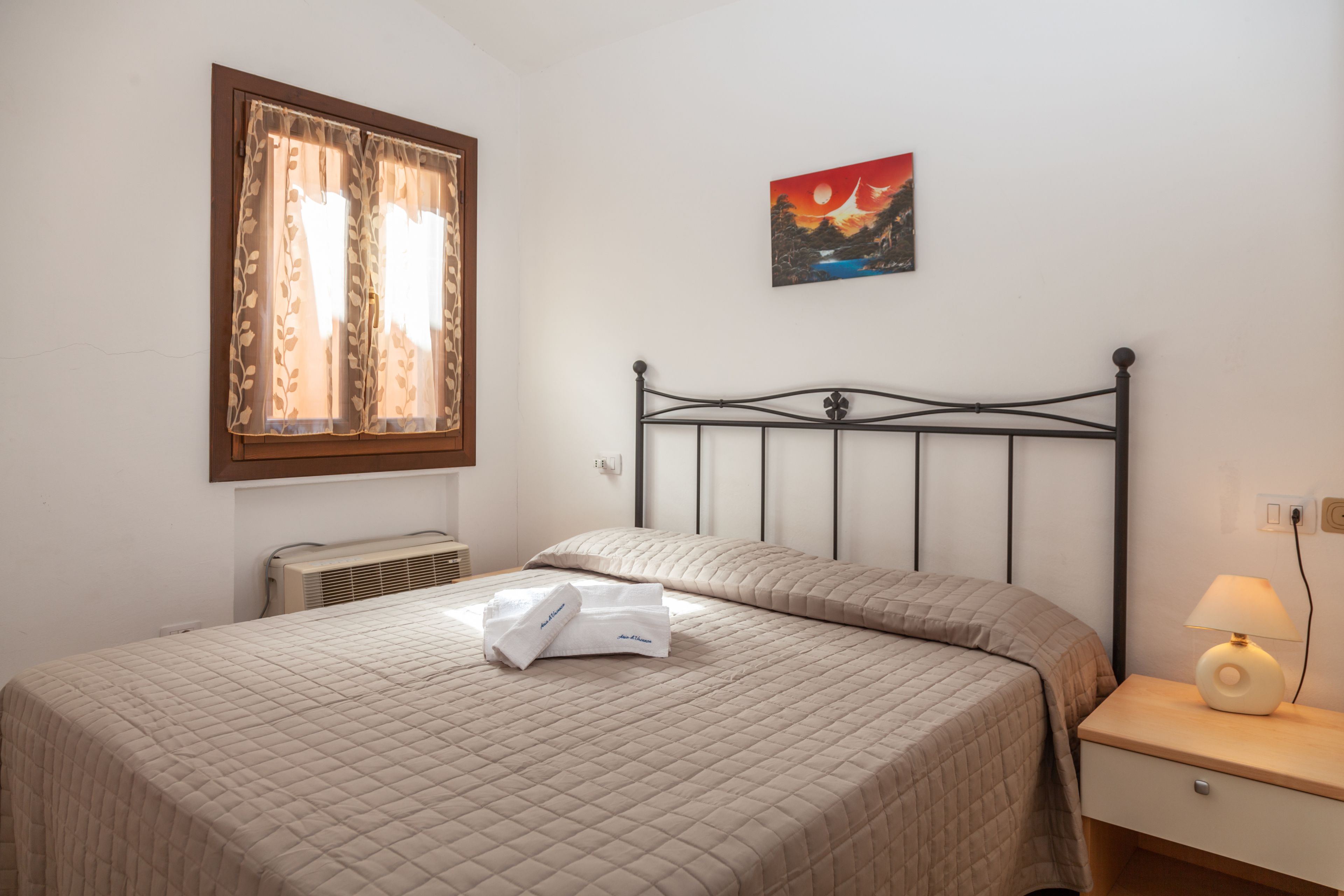 Quarto com cama, mesa de cabeceira, luminária, janela com cortinas e um quadro na parede.