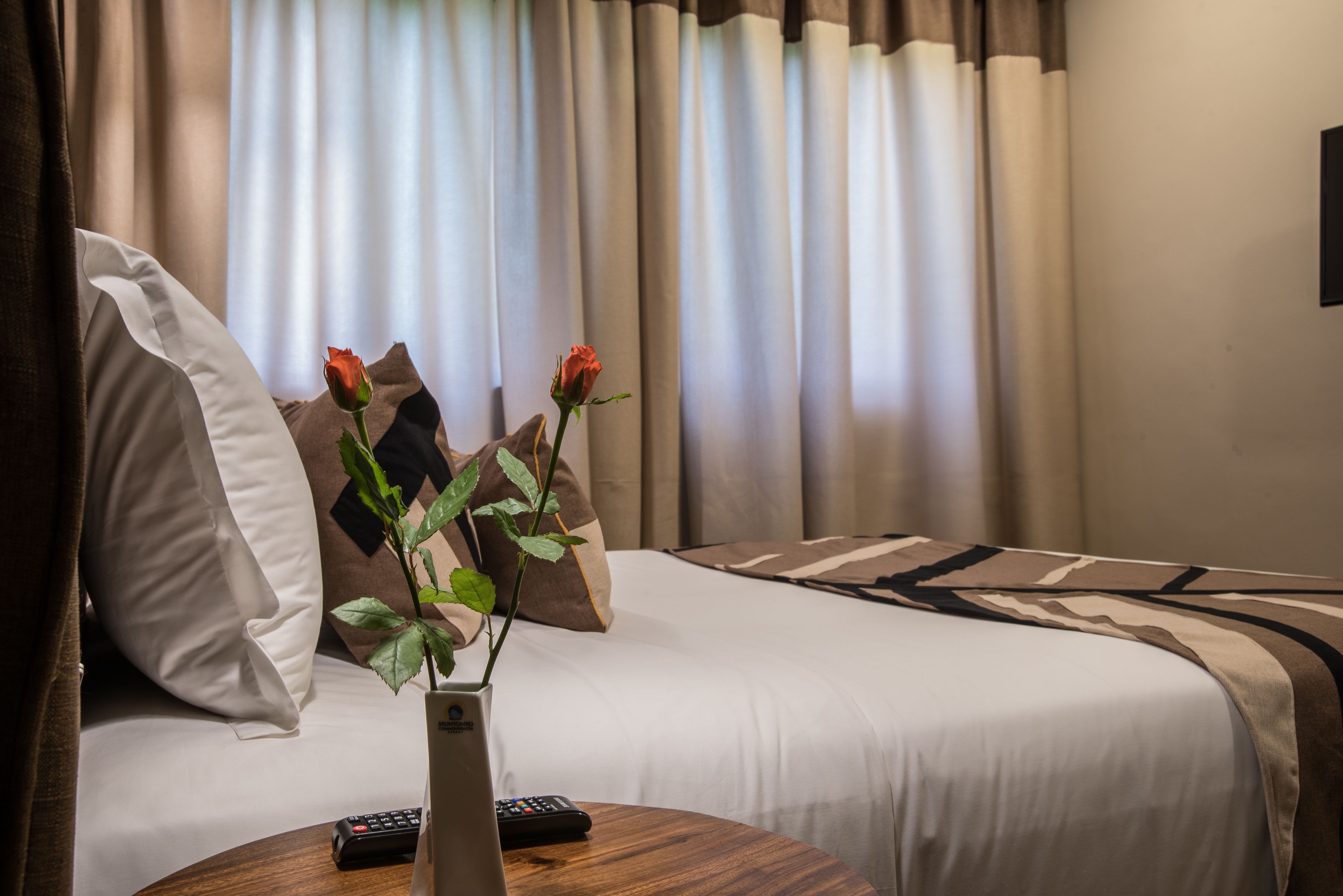 Quarto de hotel com cama, mesa de cabeceira com um vaso de flores, controle remoto e cortinas.