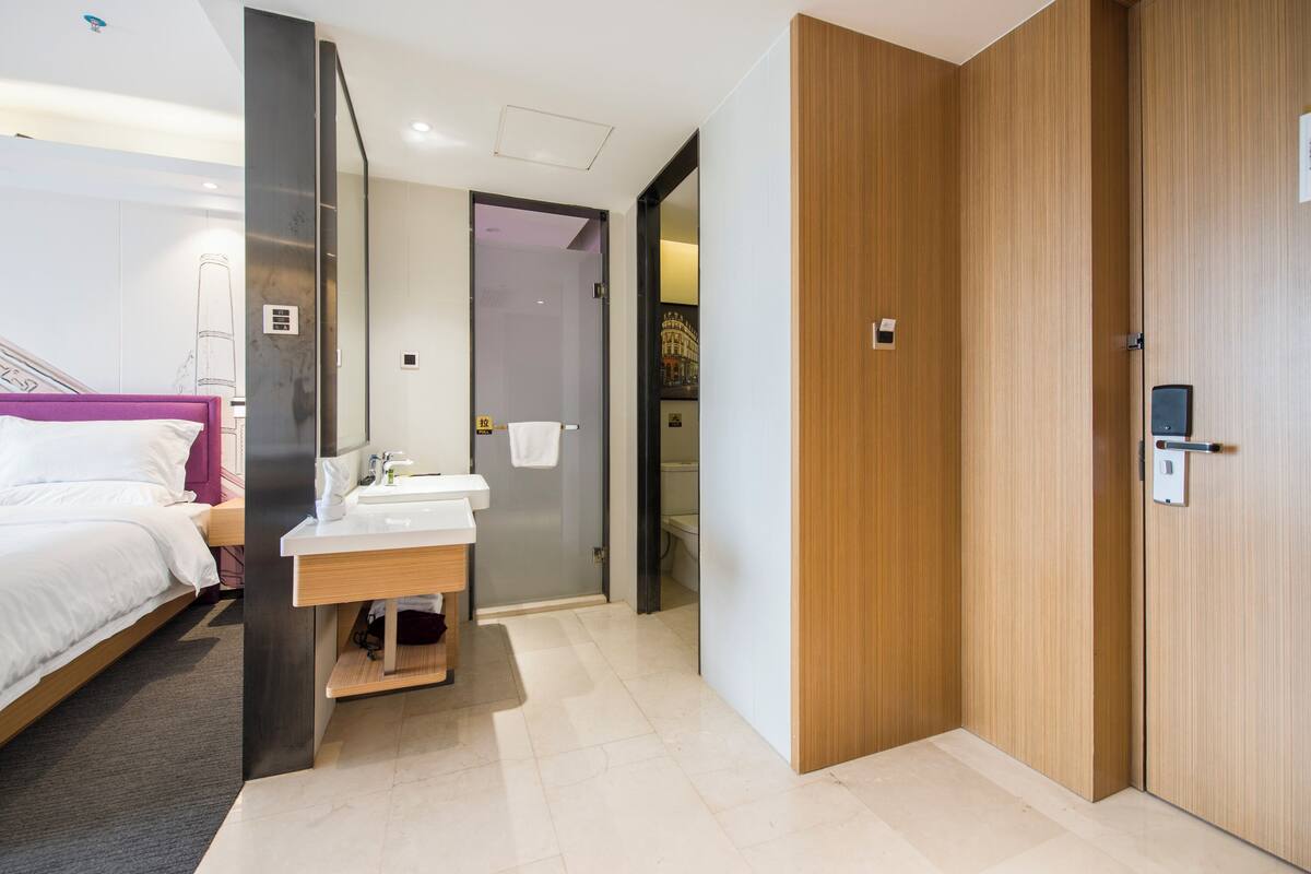 Quarto de hotel com uma cama, banheiro com pia e espelho, e uma porta de madeira.