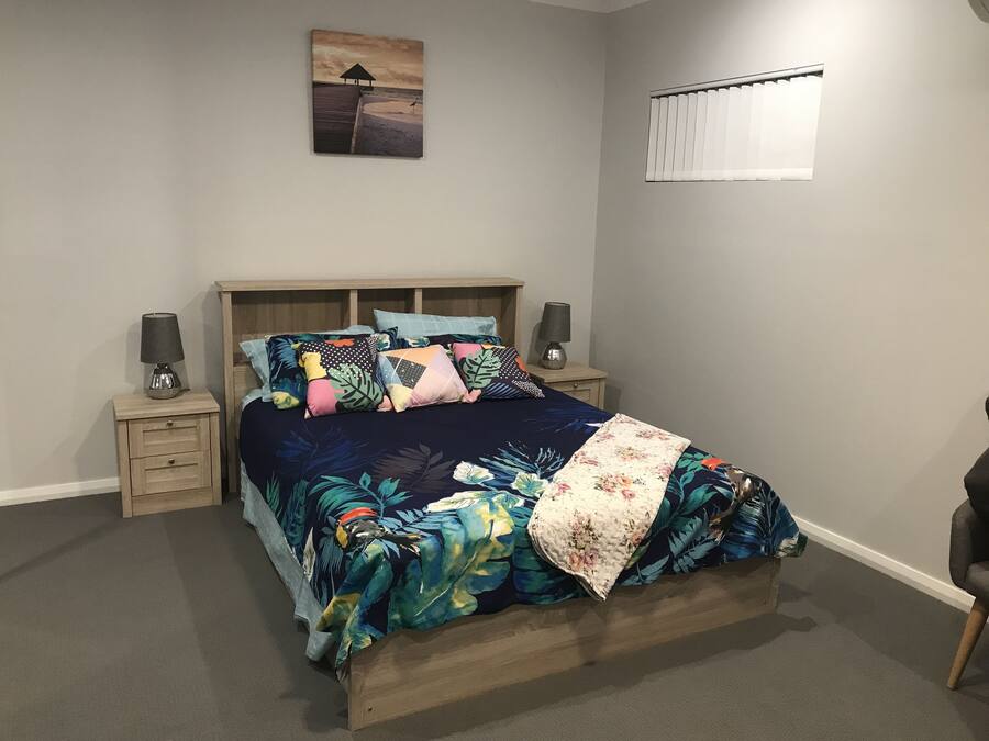 Quarto com cama de madeira, duas mesas de cabeceira com abajures, uma obra de arte na parede, janela com persianas e piso coberto por carpete.