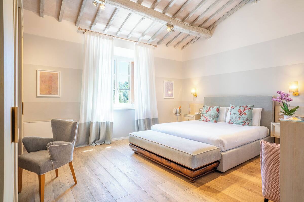 Quarto com cama, cadeira, mesa de cabeceira e janela com cortinas.