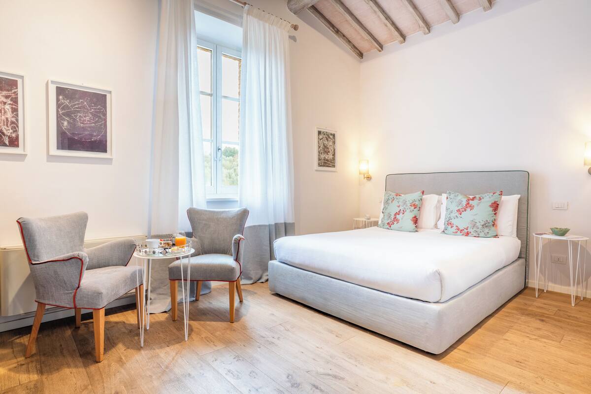 Quarto com cama, duas cadeiras, uma mesinha e obras de arte nas paredes.