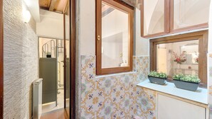 Um corredor estreito com azulejos estampados, uma porta de madeira e uma janela com plantas em vasos.