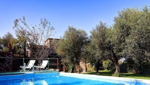 Piscina com duas espreguiçadeiras brancas, rodeada por oliveiras e um muro de pedra.