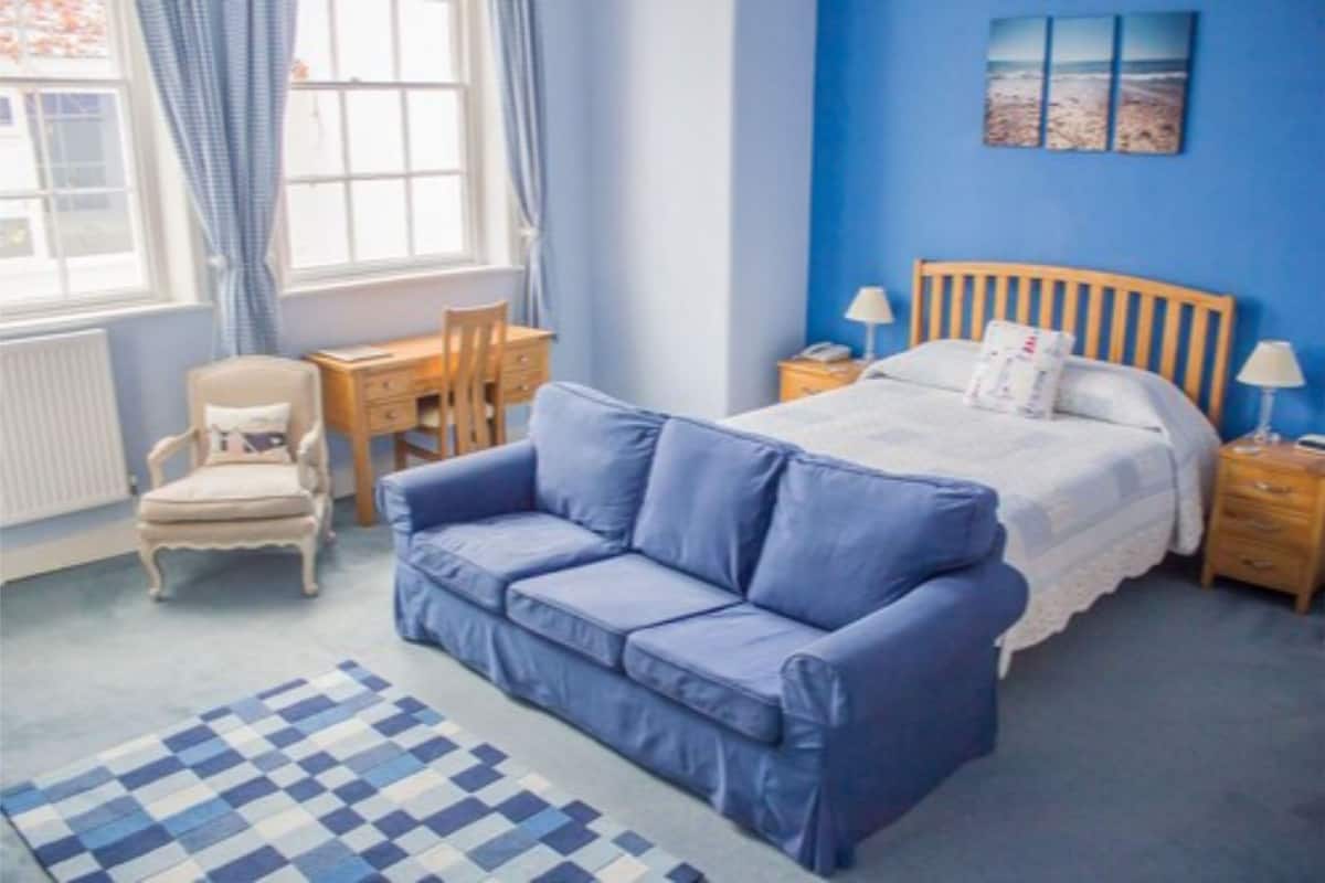 Quarto com cama, sofá azul, cadeira, mesa de cabeceira de madeira e janela com cortinas.