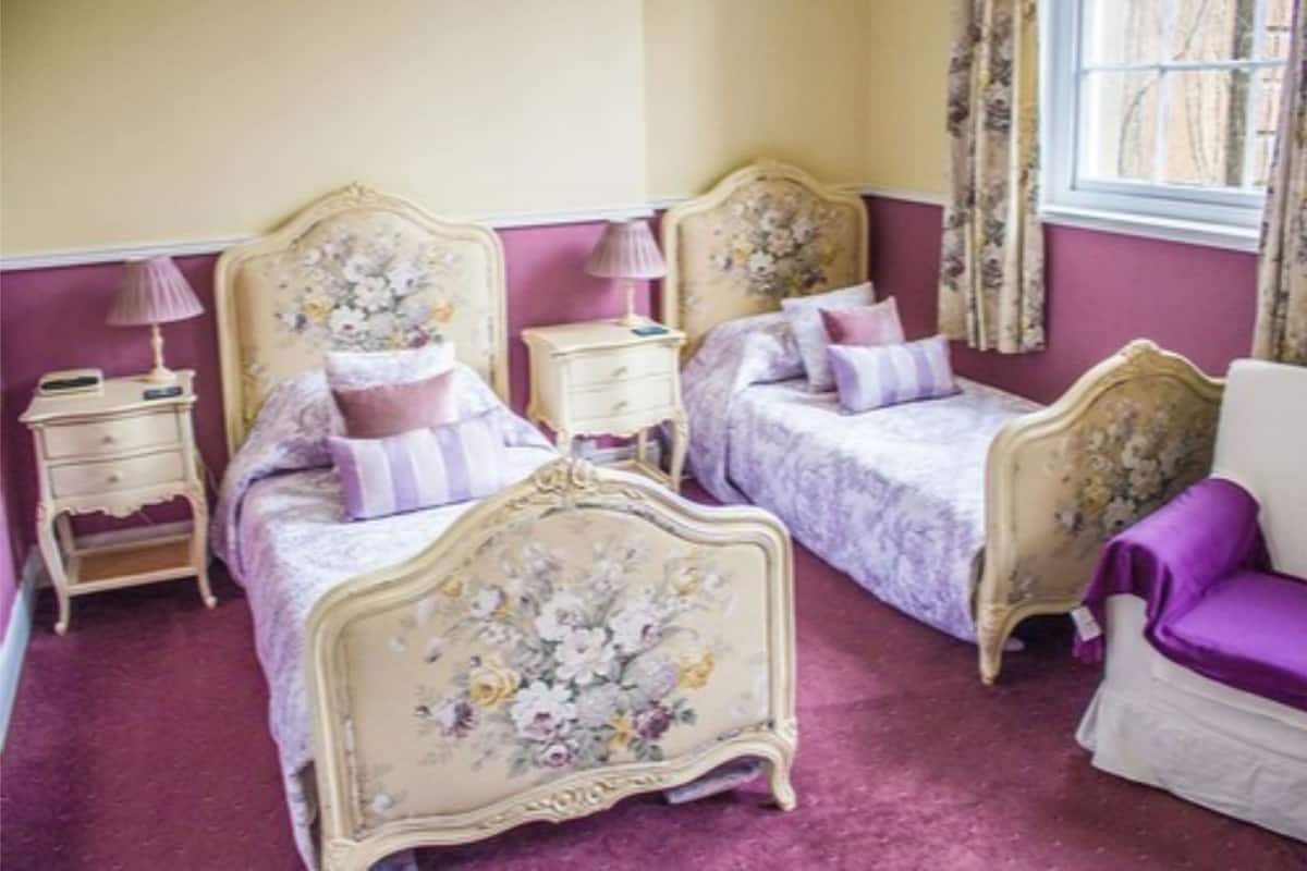 Quarto com duas camas, uma mesa de cabeceira, uma cadeira e uma janela com cortinas.