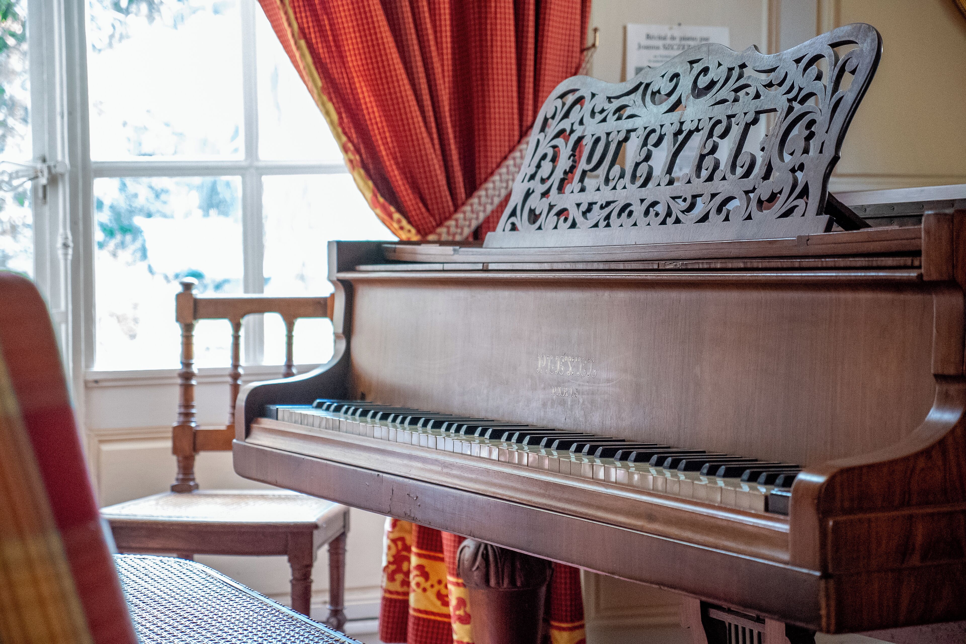Um piano de cauda em um quarto com uma cadeira de madeira e uma janela com cortinas vermelhas.