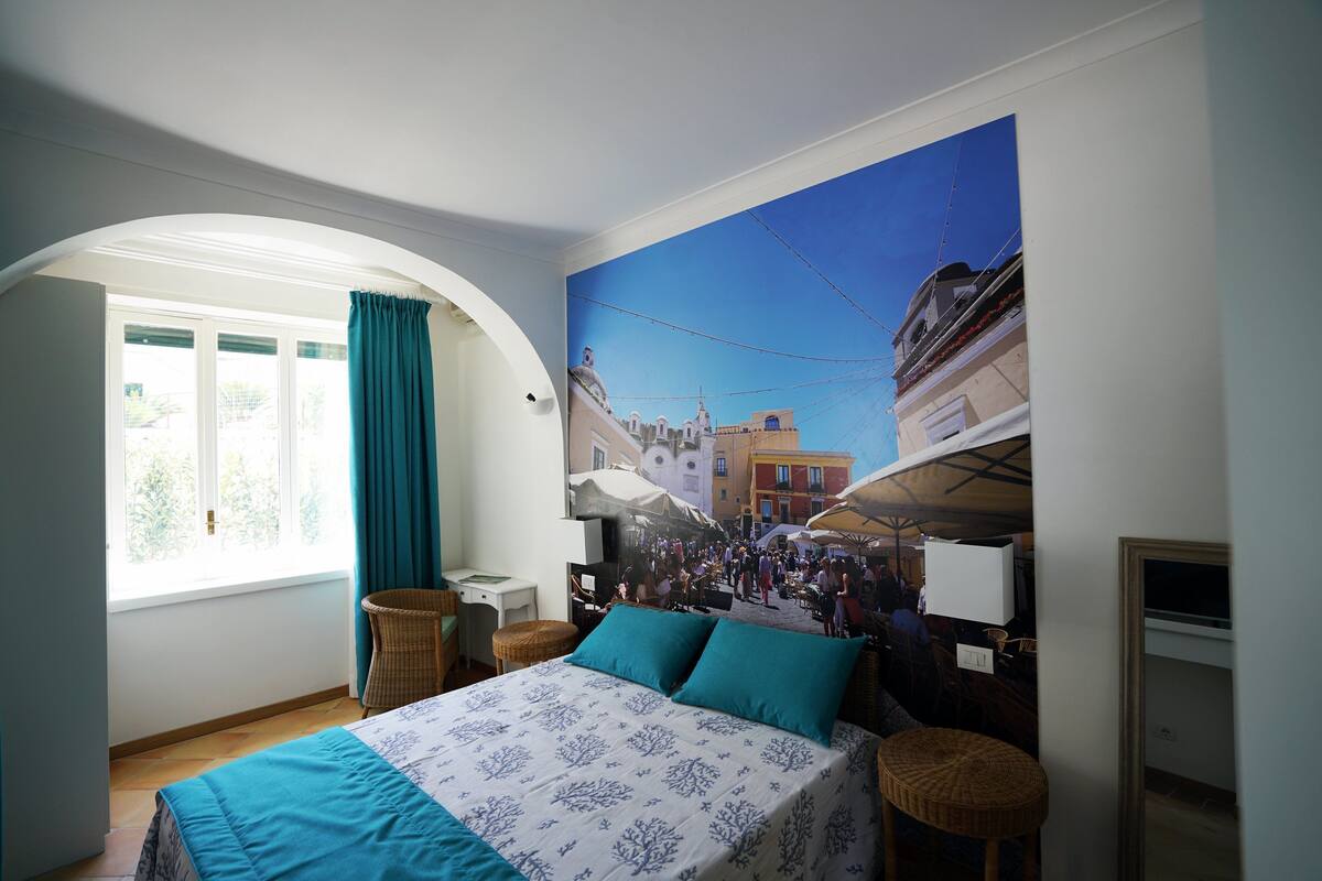 Quarto com uma cama grande, janela com cortinas, uma cadeira de vime e um mural com uma cena de rua.