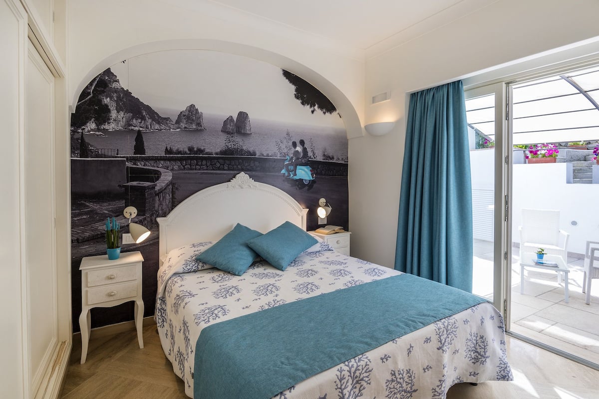 Quarto com cama, mesas de cabeceira, um mural e vista através de uma porta de vidro.