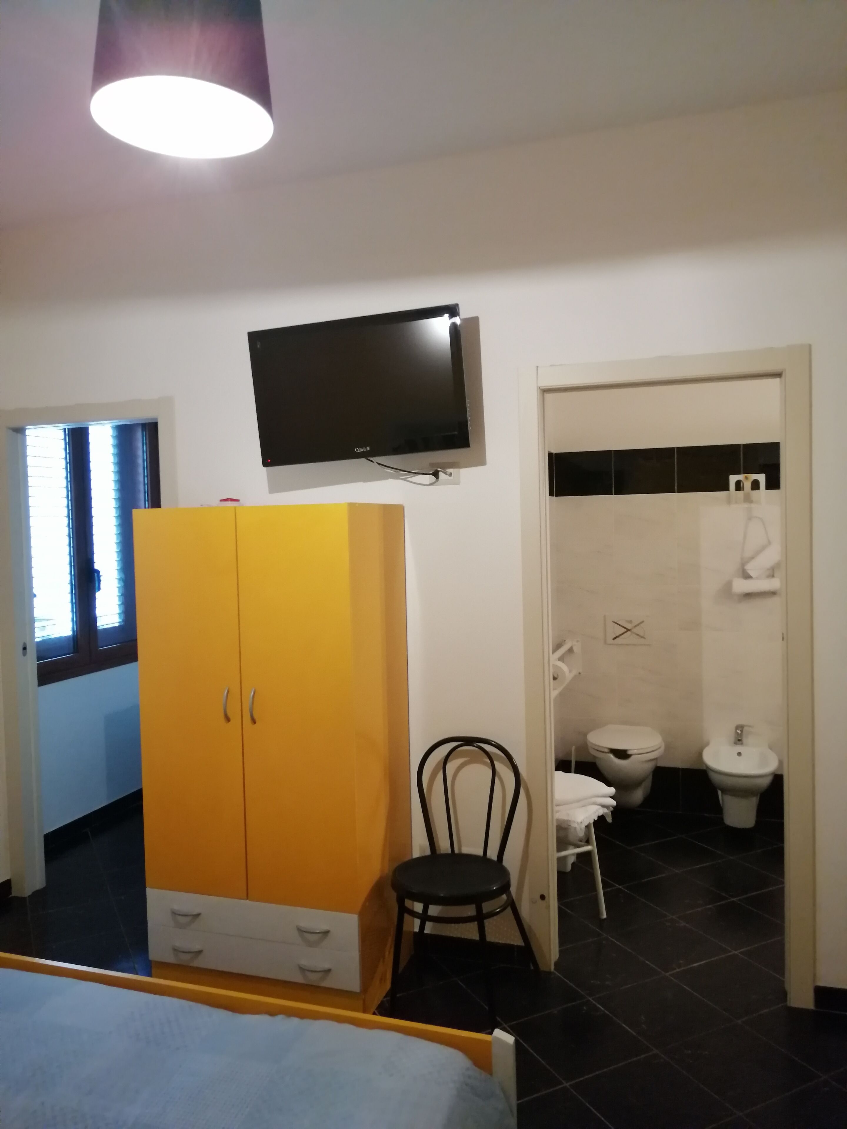 Quarto com cama, guarda-roupa amarelo, televisão na parede e banheiro com vaso sanitário, bidê e porta-toalhas.