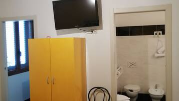 Quarto com cama, guarda-roupa amarelo, televisão na parede e banheiro com vaso sanitário, bidê e porta-toalhas.