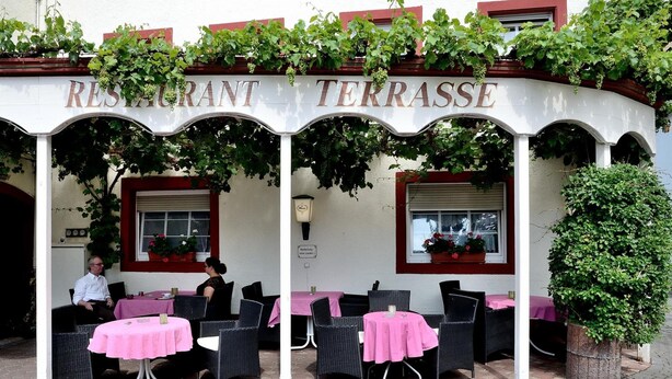 A restaurant terrace with outdoor seating, covered by a pergola with vines, and a sign reading "RESTAURANT TERRASSE".