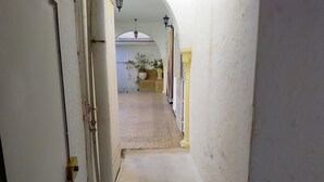 Um corredor estreito com paredes brancas e chão de azulejos, que dá para um quarto com teto estampado e luminária pendente.