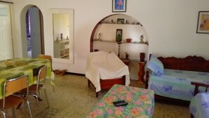 Quarto com mesa de jantar, duas cadeiras, cama com colcha florida, uma pequena mesa com controle remoto e um espelho.