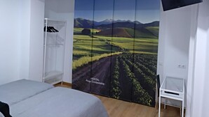 Quarto com cama, uma mesinha, guarda-roupa e uma janela grande com vista panorâmica para um campo e montanhas.