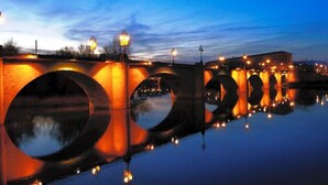 Uma ponte de pedra com arcos sobre águas calmas, iluminada por postes de luz.