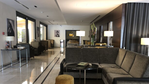 Um saguão moderno de hotel com área de estar, balcão de recepção e uma grande janela que proporciona iluminação natural.