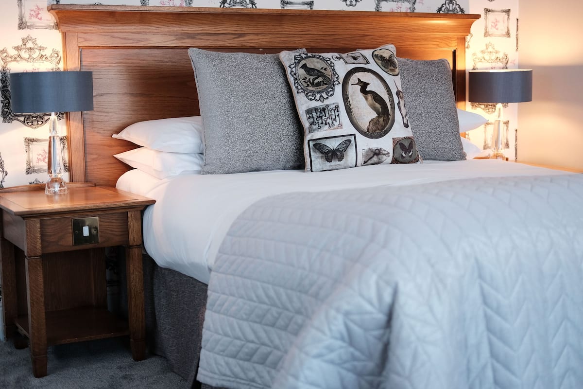 Cama bem arrumada com cobre branca acolchoada, cabeceira de madeira e almofadas decorativas com diferentes desenhos. Há duas mesas de cabeceira com luminárias, uma de cada lado da cama.