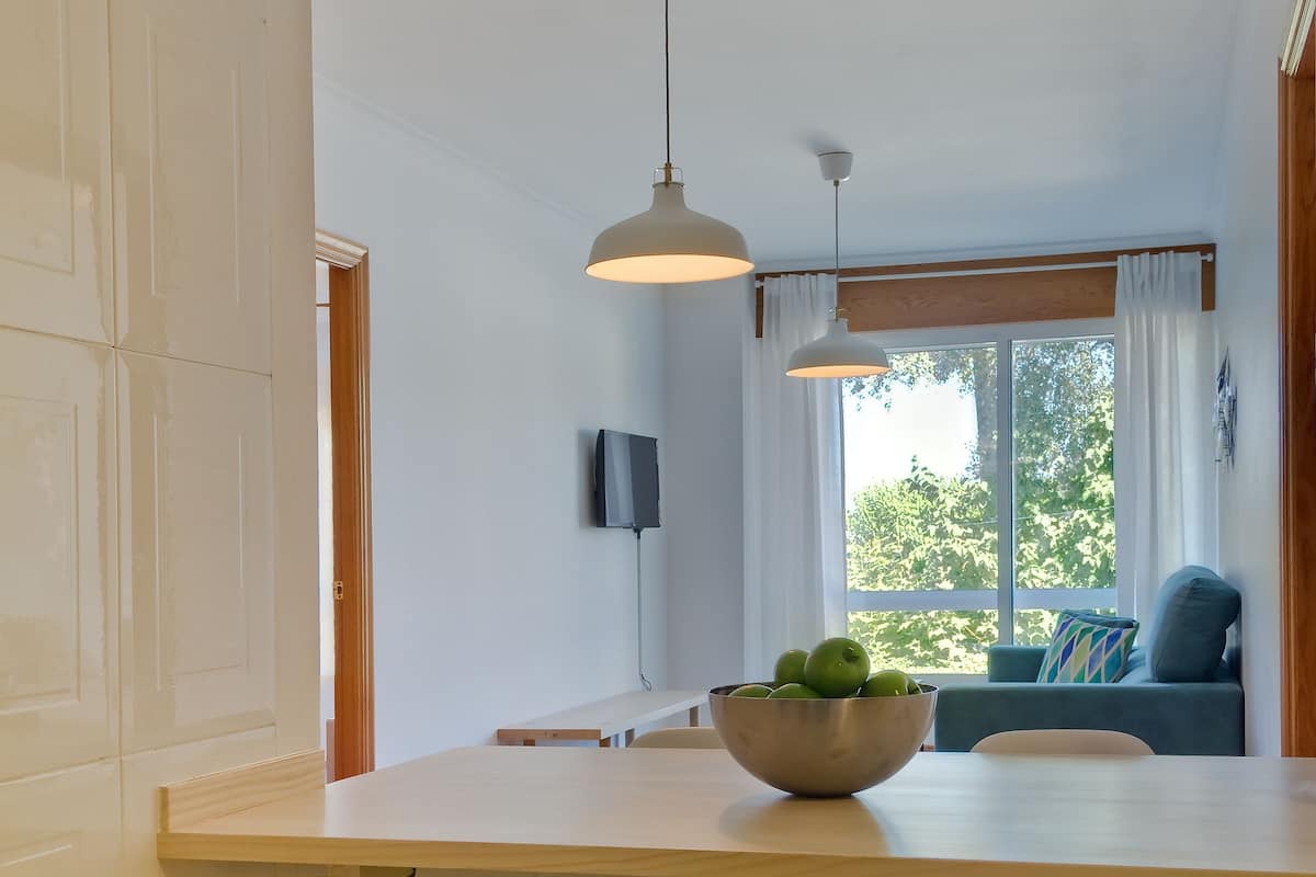 Uma cozinha moderna com bancada de madeira, duas cadeiras brancas, uma tigela de frutas, uma luminária pendente e vista para o exterior através de uma janela.