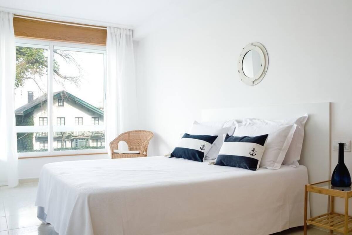 Quarto bem arrumado com cama, uma cadeira, mesa de cabeceira e janela com vista para prédios.