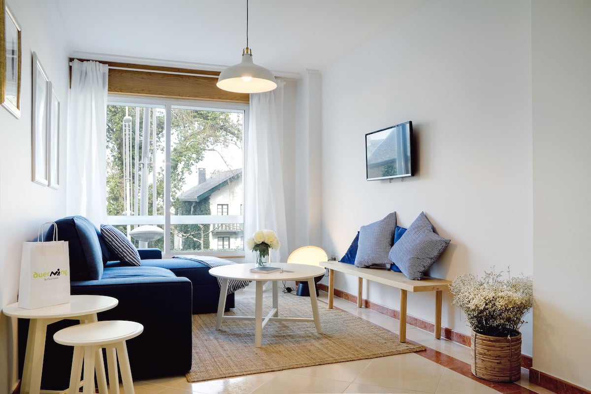Uma sala de estar moderna com um sofá, uma mesa de centro, uma TV fixada na parede e uma janela com cortinas.