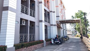 Um edifício residencial com vários andares, varandas, aparelhos de ar condicionado e uma área de estacionamento coberta com scooters estacionadas.