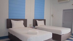 Quarto de hotel com duas camas, cada uma com cabeceira marrom e roupa de cama branca. Há painéis azuis e brancos na parede, uma porta e um aparelho de ar condicionado.
