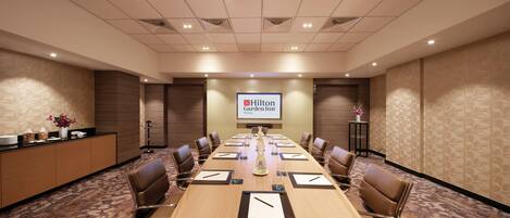 Sala de conferências com uma mesa comprida, cadeiras, um púlpito e uma tela exibindo o logotipo do Hilton Garden Inn.