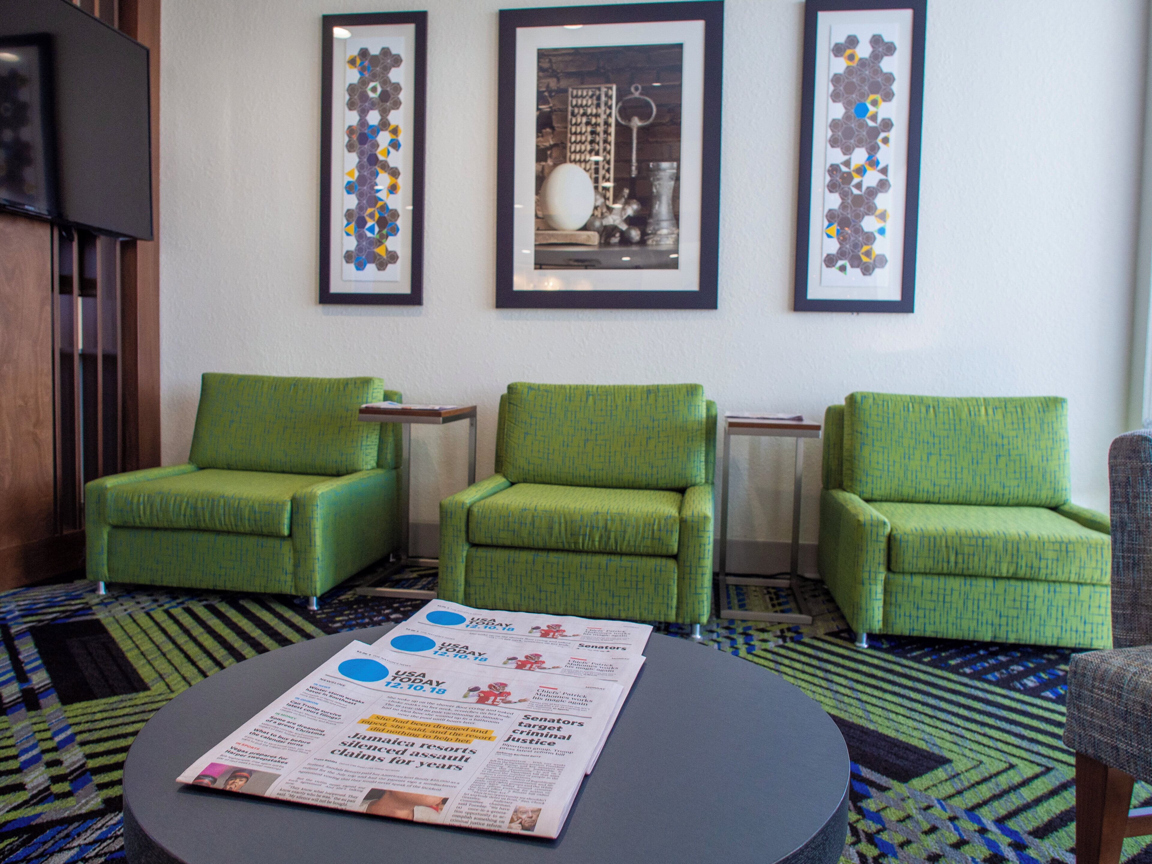 Um lobby de hotel com poltronas verdes, uma mesa redonda com um jornal e obras de arte emolduradas na parede.