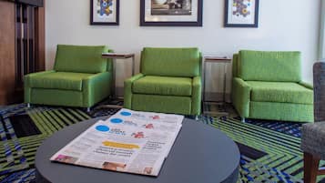 Um lobby de hotel com poltronas verdes, uma mesa redonda com um jornal e obras de arte emolduradas na parede.