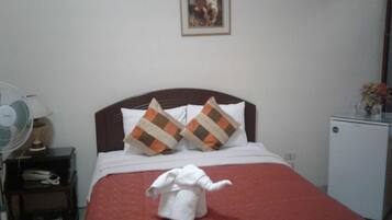 Quarto de hotel com cama, almofadas, ventilador, pequeno frigorífico e toalha em forma de elefante.