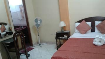 Quarto de hotel com cama, escrivaninha, cadeira, ventilador e mesa de cabeceira.