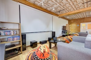Uma sala de estar aconchegante com teto de madeira, tela de projetor, sofá, mesa de centro e estante repleta de livros e DVDs.