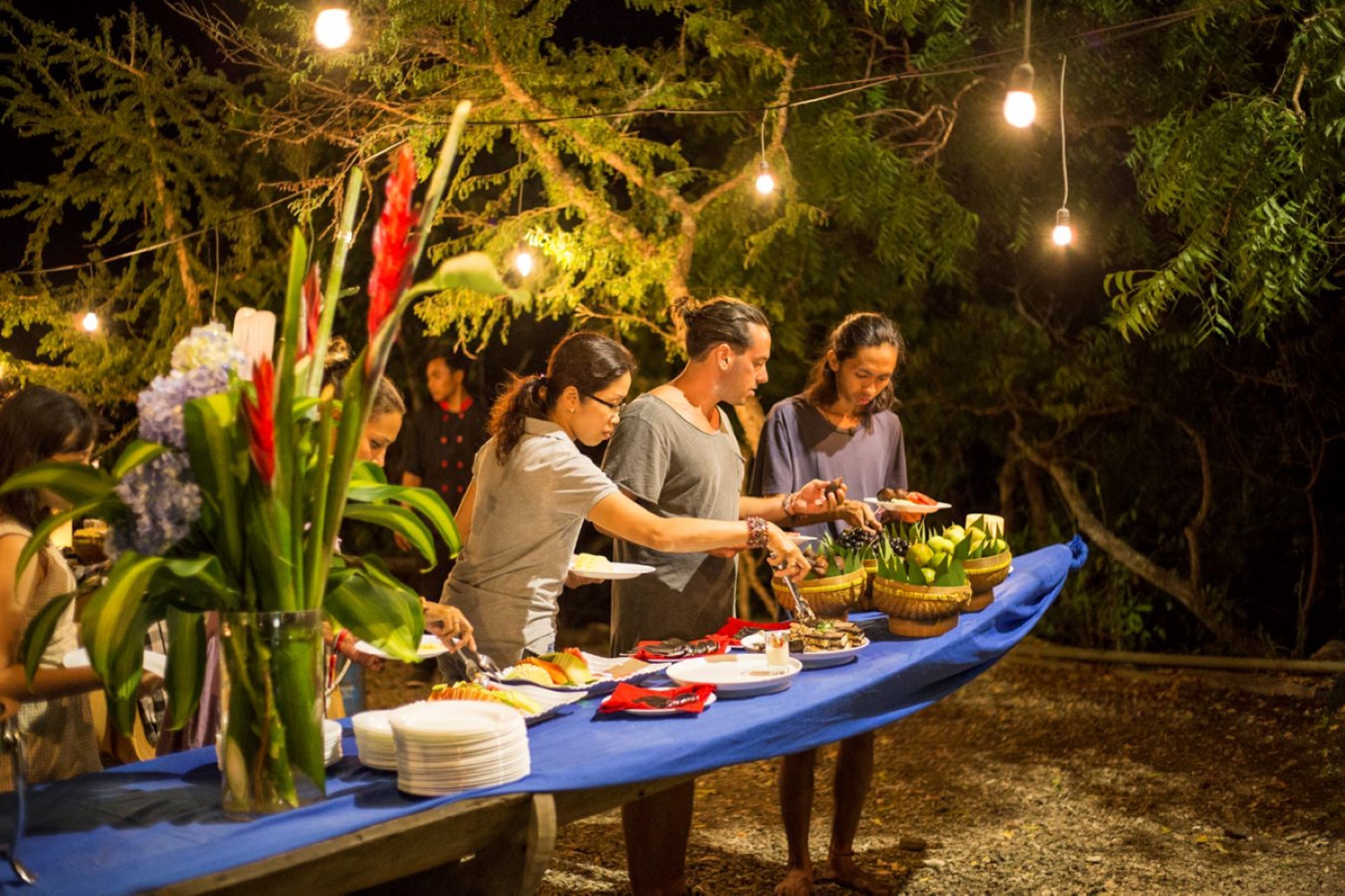 Uma mesa de buffet com vários pratos e uma toalha de mesa azul, rodeada por convidados.
