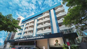 Um hotel de vários andares com uma fachada azul e branca destacada, várias varandas e um estacionamento com diversos carros.