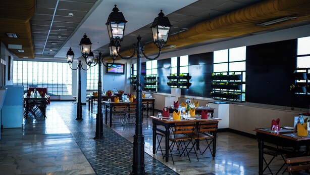 Um restaurante espaçoso com pé direito alto, grandes janelas e um lustre moderno. As mesas estão dispostas com guardanapos amarelos e vermelhos, e há plantas em vasos nos parapeitos das janelas.