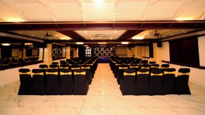 Uma sala de conferências com fileiras de cadeiras pretas com fitas amarelas, piso de mármore e teto com iluminação embutida e um ventilador.
