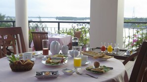 Mesa de café da manhã com diversos alimentos e bebidas, montada em uma varanda com vista para o mar.