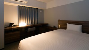 Quarto de hotel com uma cama grande, uma escrivaninha com abajur, uma cadeira e uma janela com cortinas.