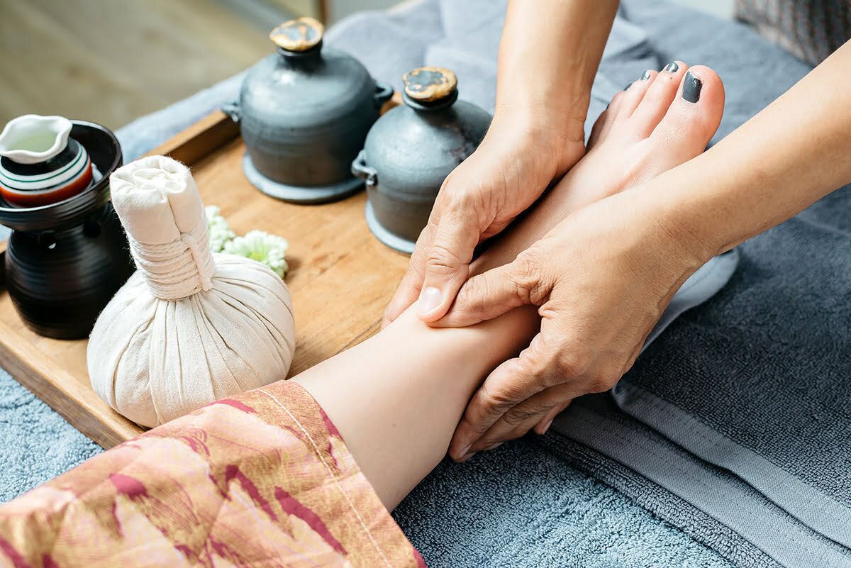 Massagem nos pés com bandeja de madeira contendo um massageador para pés, uma pedra branca e dois recipientes pretos.