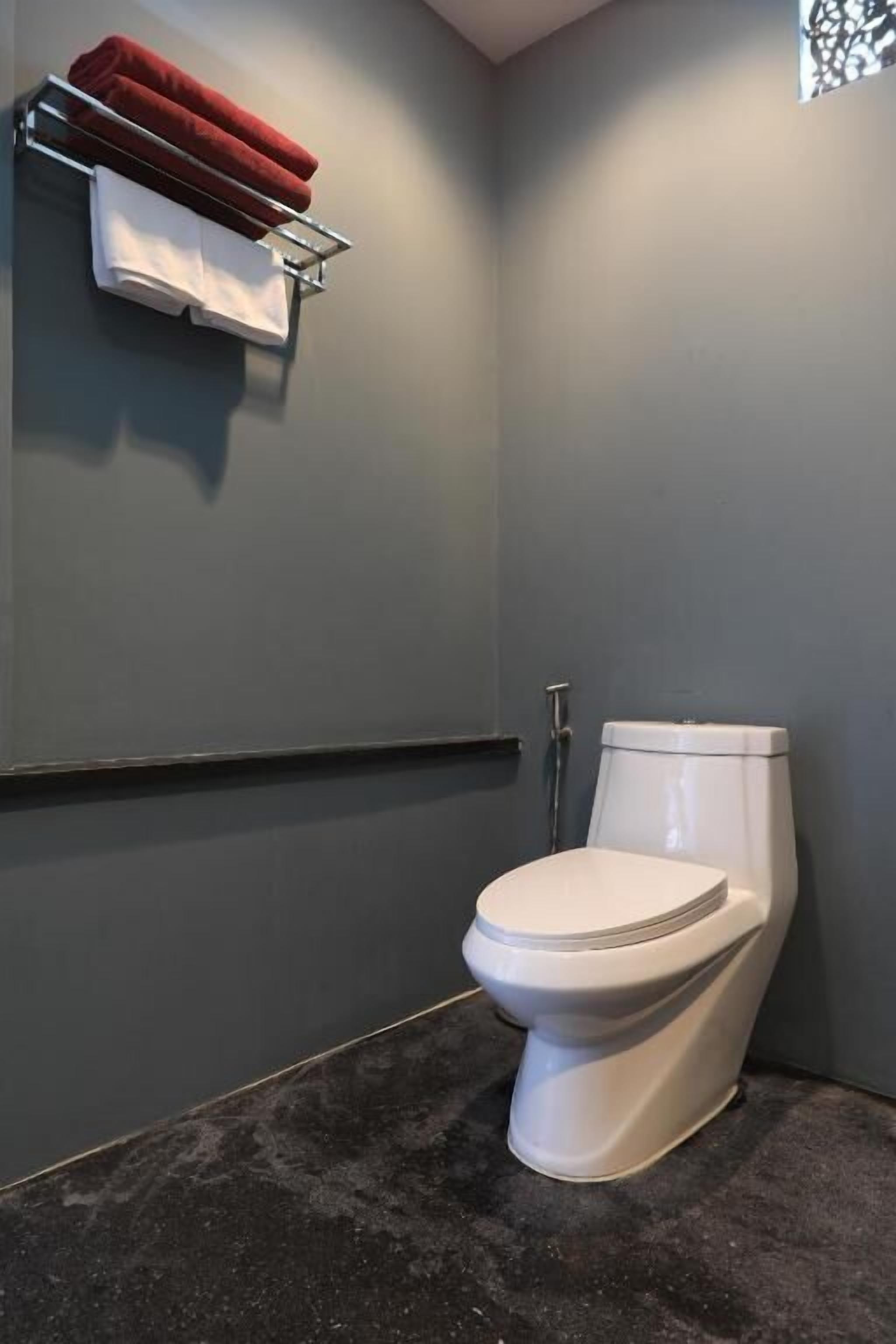 Casa de banho com uma sanita branca, um porta-toalhas com toalhas vermelhas e brancas, e um chão escuro.