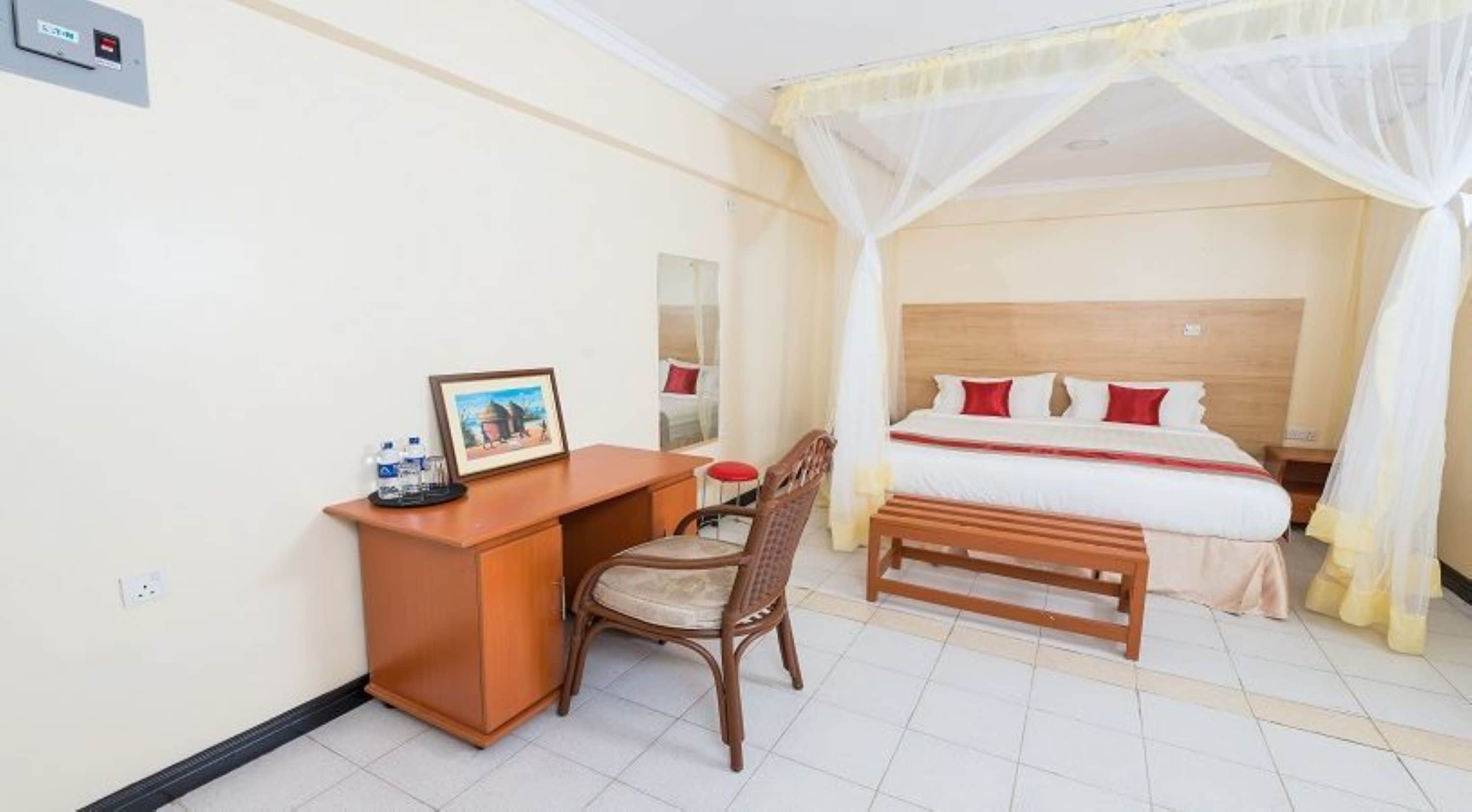 Quarto de hotel com cama, escrivaninha, cadeira, mesa de cabeceira, moldura e janela com cortinas.