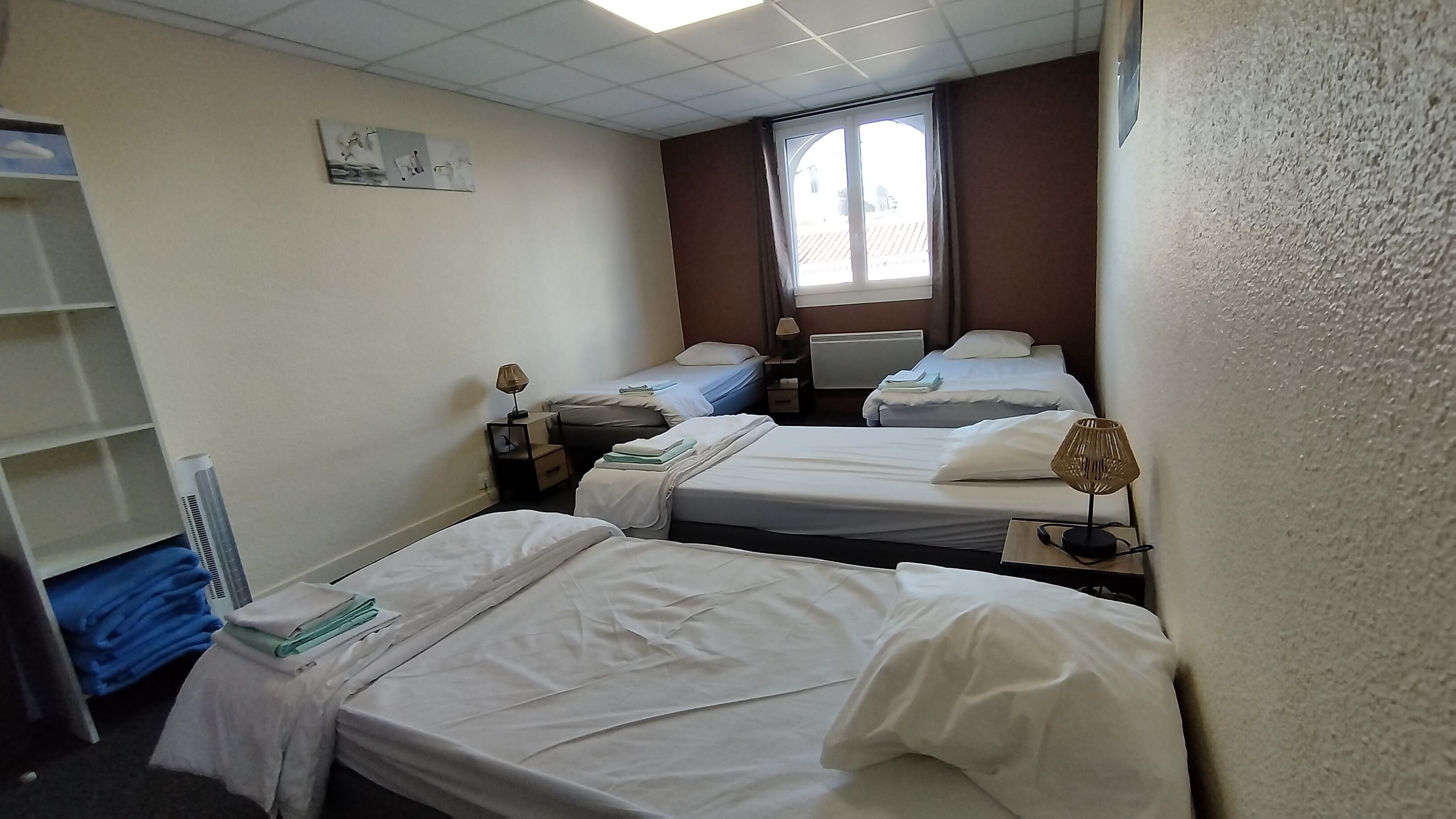 Quarto de hotel com duas camas, uma janela, um radiador, uma luminária e um quadro pendurado na parede.
