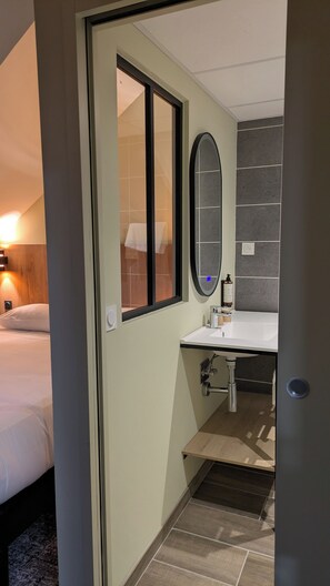Quarto de hotel pequeno com cama, banheiro com pia e espelho, e vista do quarto pela janela.