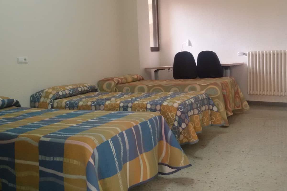 Quarto com três camas de solteiro, cada uma com colcha estampada. Ao fundo, há uma escrivaninha com duas cadeiras e um radiador na parede.