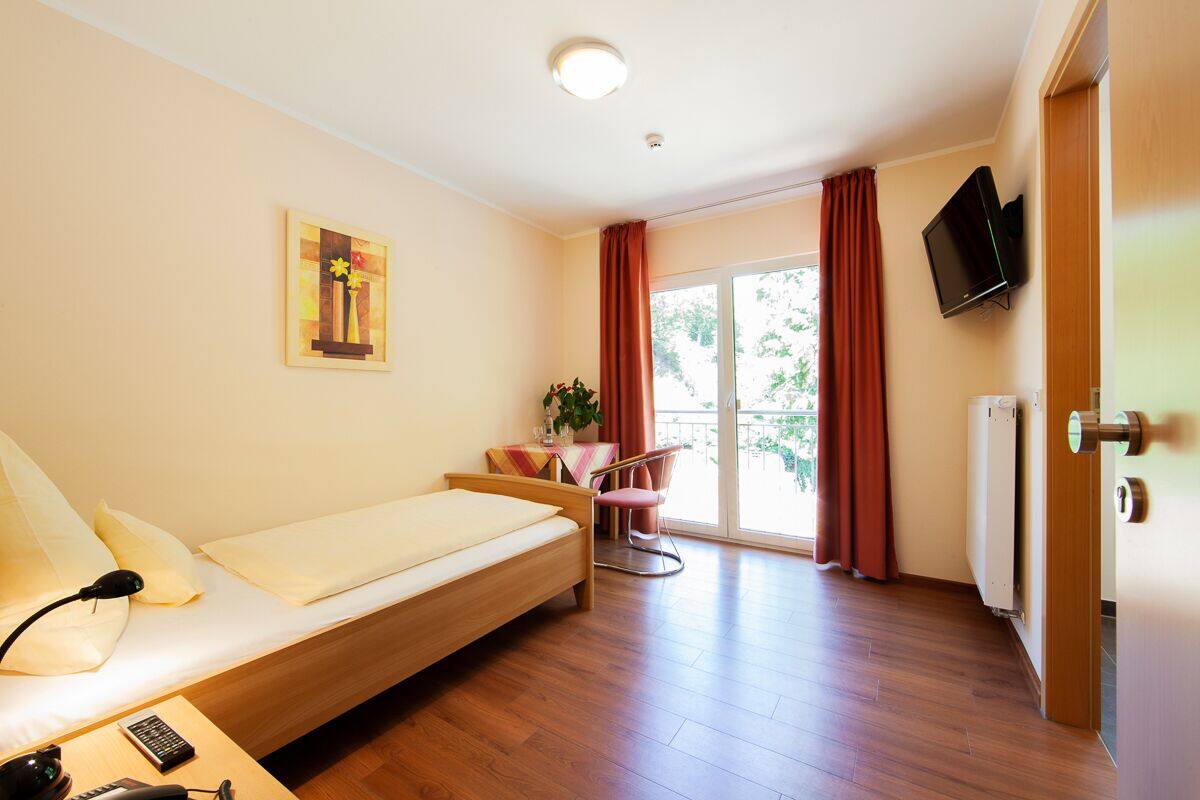 Quarto de hotel com uma cama, uma escrivaninha com cadeira, televisão fixada na parede e janela com cortinas.