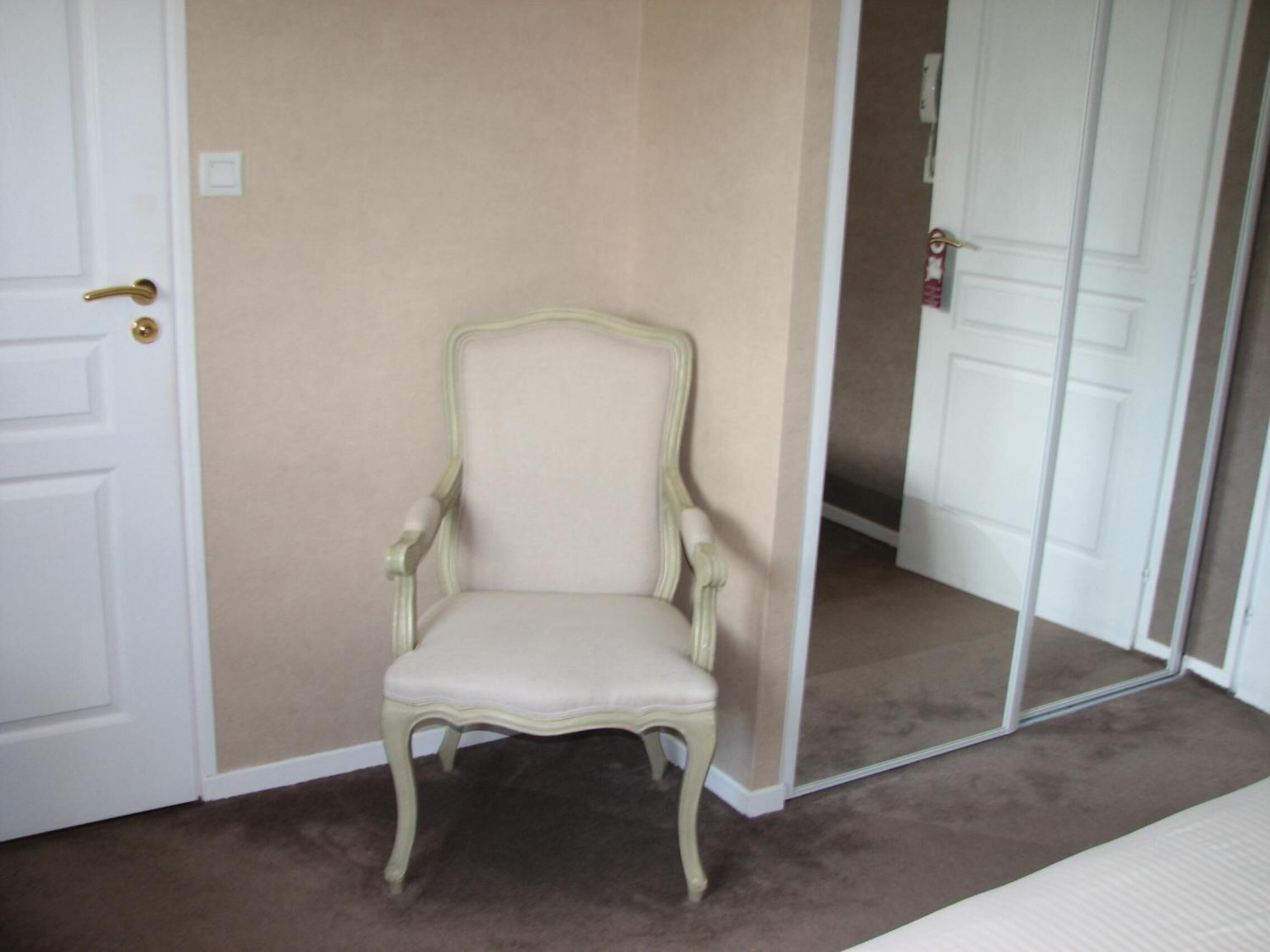 Quarto com uma única cadeira em frente a uma parede bege, uma porta branca e um espelho refletindo parte do espaço.